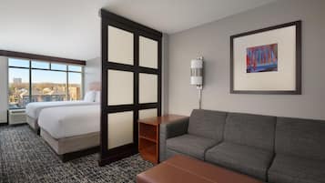 Quarto de hotel com duas camas, um sofá cinzento, um banquinho marrom, um quadro na parede e uma janela com vista para a cidade.