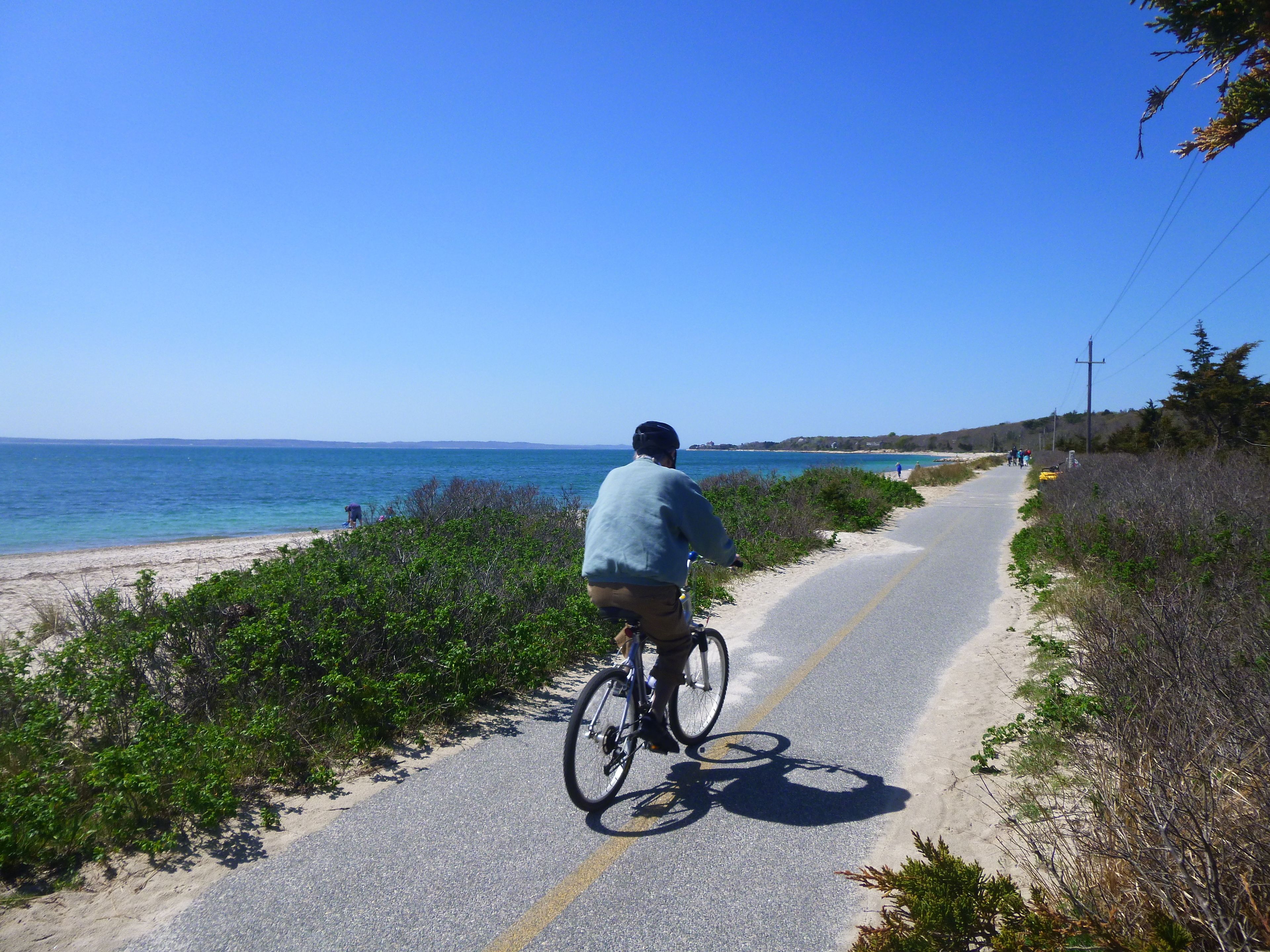 Um caminho pavimentado ao longo da praia, com um ciclista e algumas pessoas à distância.