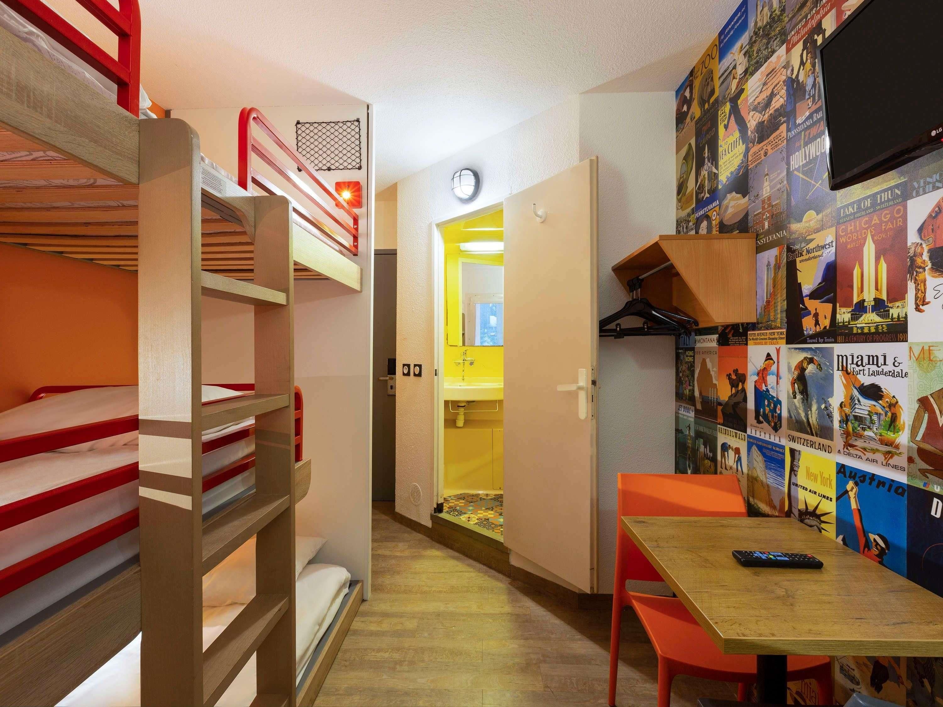Quarto com beliches, uma escrivaninha com cadeira, televisão e parede decorada com cartazes de viagens.