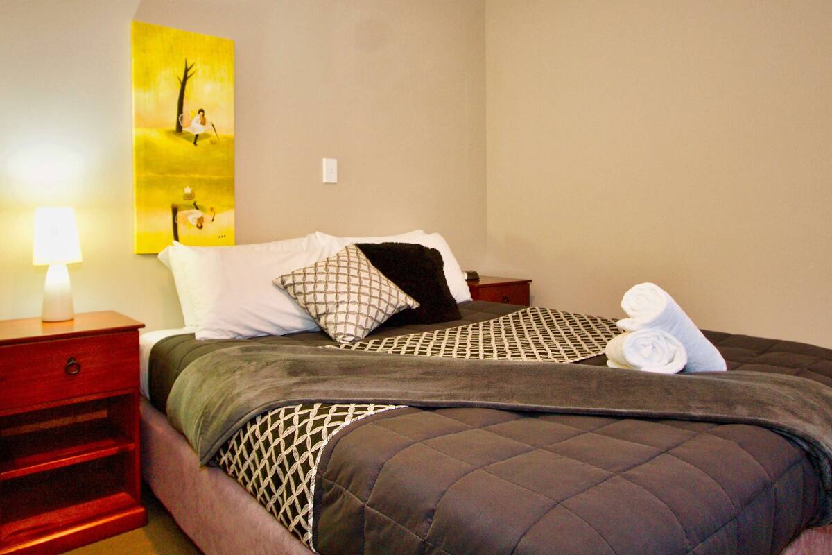 Cama bem arrumada com manta acolchoada, duas almofadas e toalhas dobradas. Há uma mesa de cabeceira com abajur e uma gavetinha. Um quadro está pendurado na parede acima da cama.