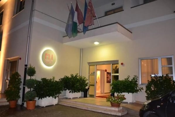Entrada de hotel com uma placa circular, plantas em vasos e bandeiras.