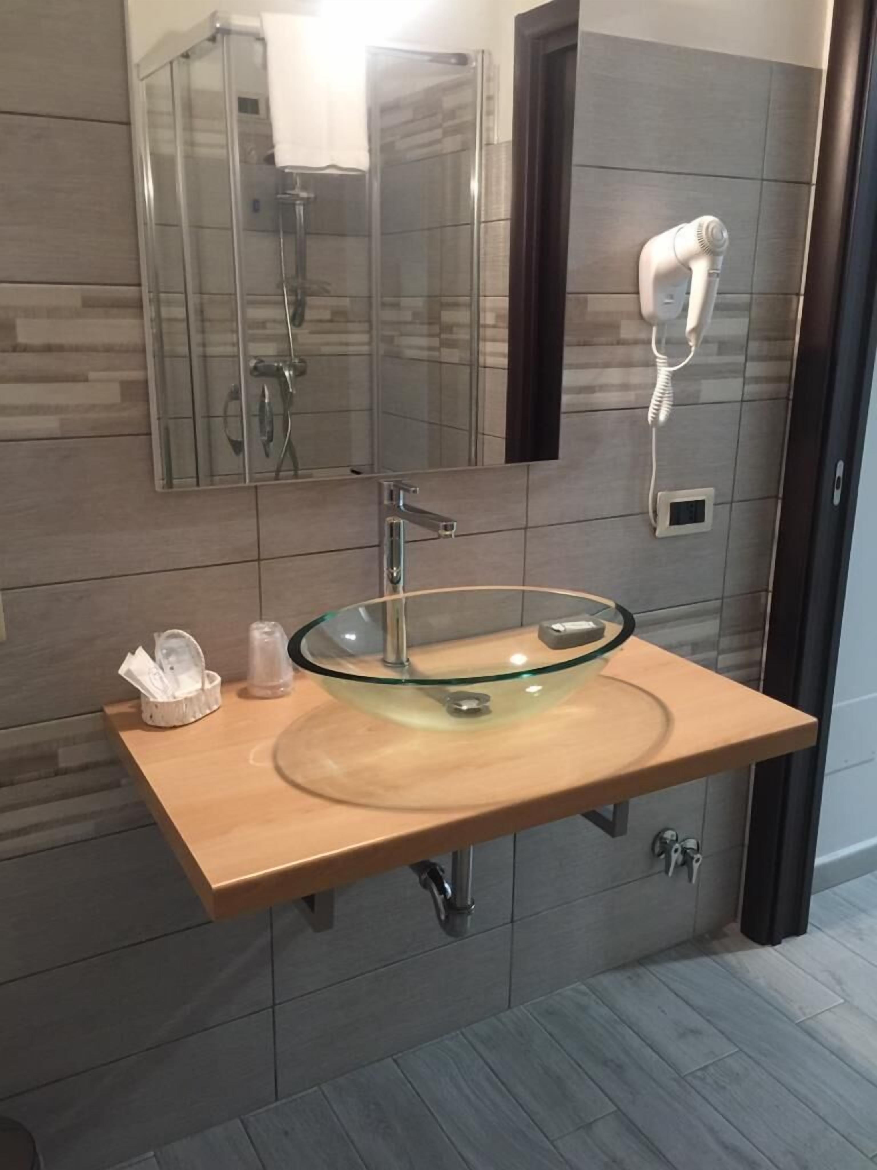 Casa de banho moderna com lavatório de vidro, chuveiro manual e secador de cabelo fixado na parede.