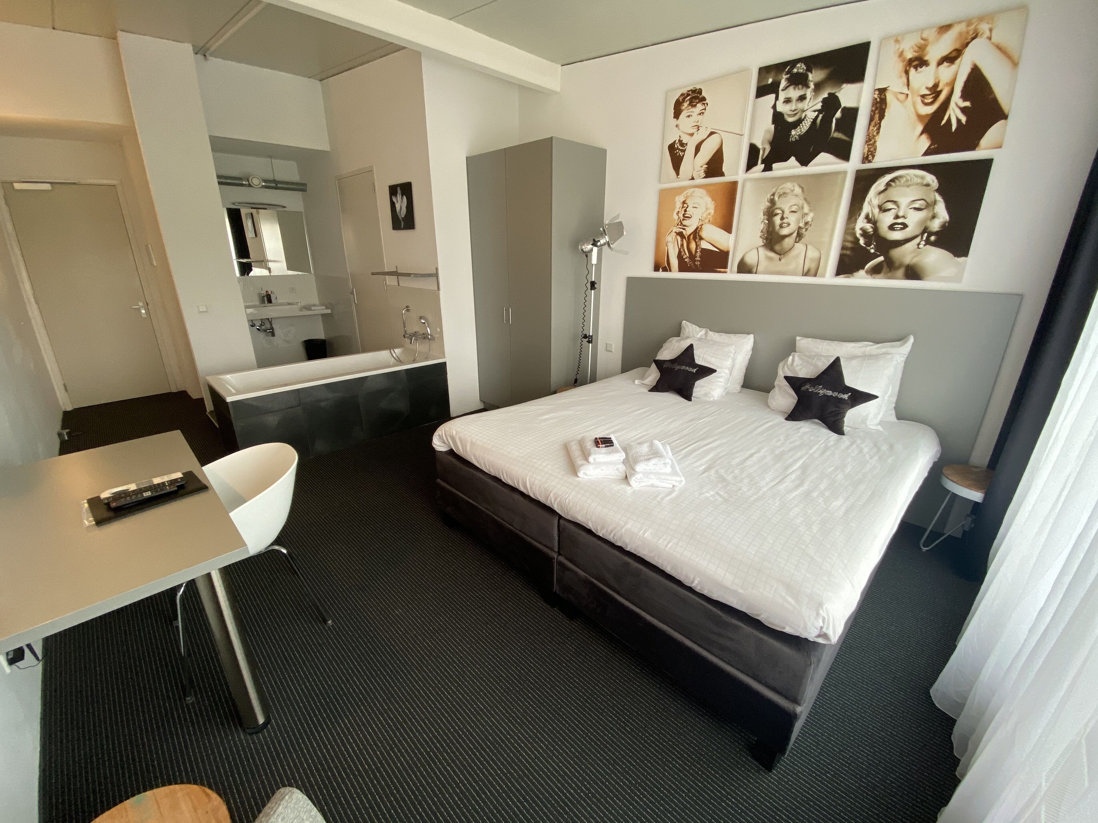 Quarto de hotel com cama, escrivaninha, cadeira, espelho e banheiro.