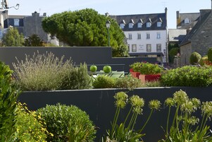 Um jardim com diversas plantas e flores, um edifício com a placa 'Hotel du Centre' e uma árvore ao fundo.