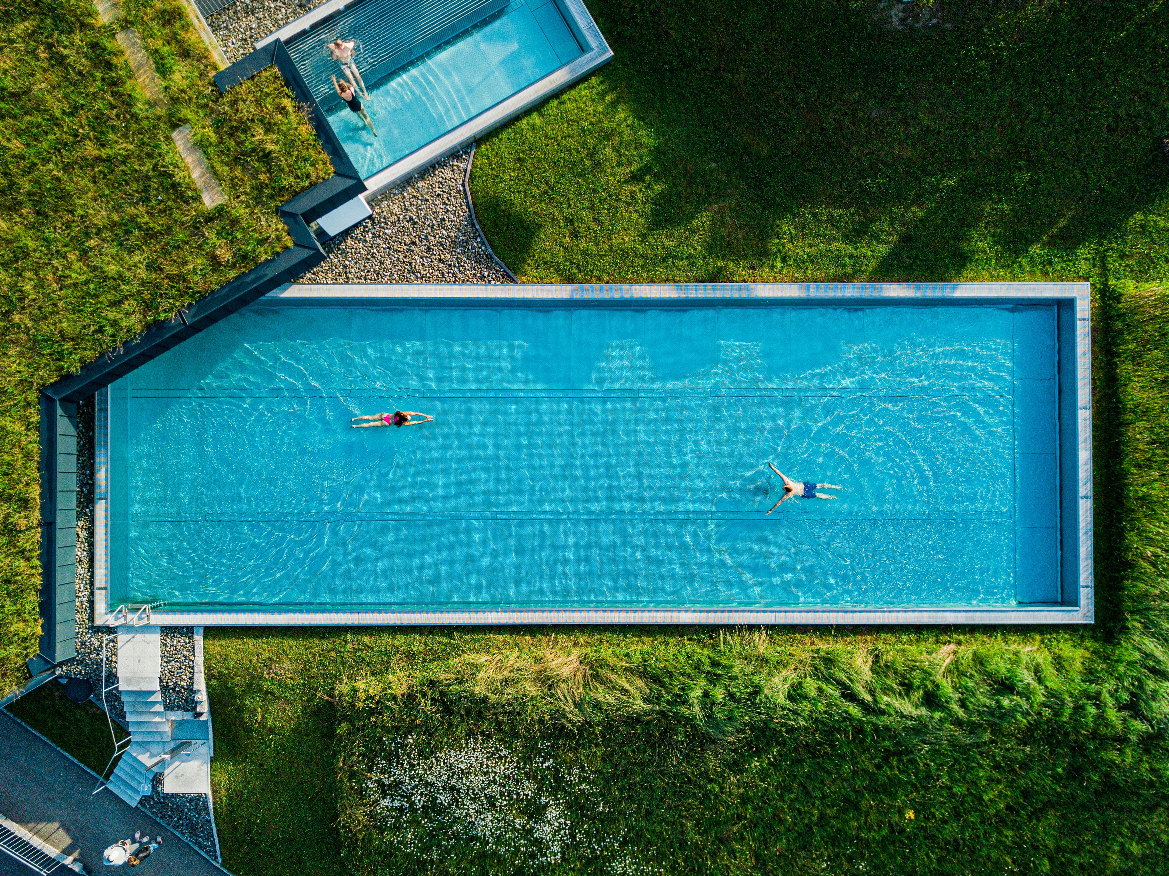 Vista aérea de uma piscina retangular com duas pessoas nadando, cercada por um gramado e um pequeno deck.