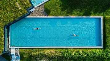Vista aérea de uma piscina retangular com duas pessoas nadando, cercada por um gramado e um pequeno deck.