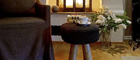 Uma sala de estar acolhedora com lareira, uma poltrona confortável, uma mesa redonda pequena com uma xícara e flores, e um enfeite pendurado na parede.