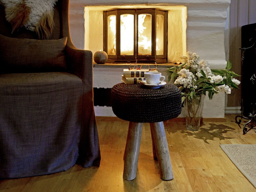 Uma sala de estar acolhedora com lareira, uma poltrona confortável, uma mesa redonda pequena com uma xícara e flores, e um enfeite pendurado na parede.