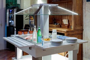 Disposição de buffet com estrutura superior em aço inoxidável, um dispensador de bebidas e diversos alimentos sobre uma mesa branca.