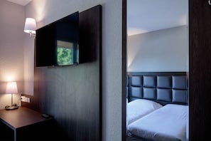 Quarto de hotel com cama, mesa de cabeceira com abajur, TV de tela plana na parede e janela com vista para o verde.