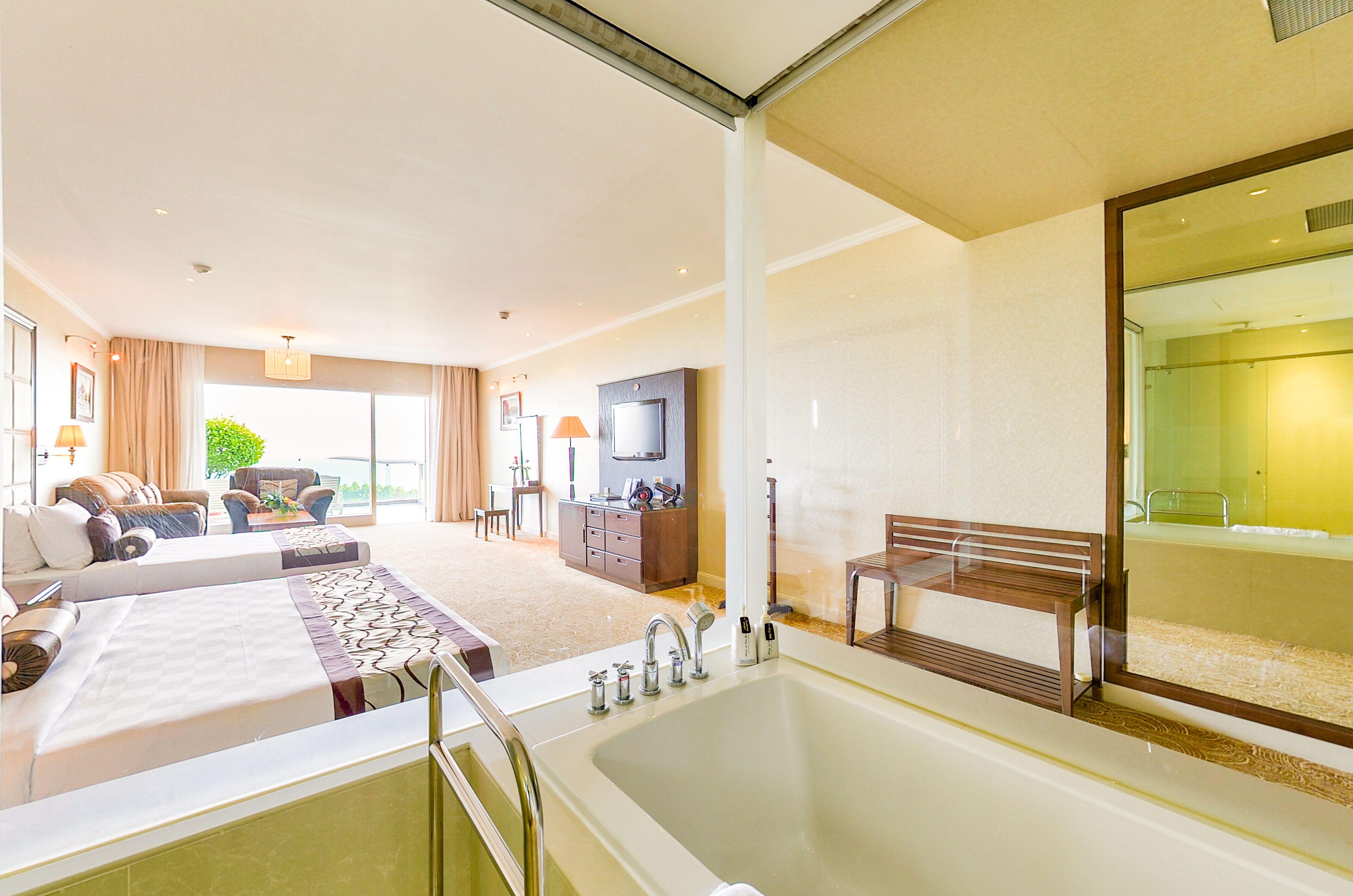 Quarto de hotel moderno com uma cama grande, área de estar com sofá e poltronas, mesa de jantar, televisão e banheiro com banheira e um espelho amplo.