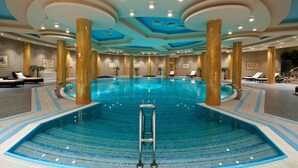 Uma grande piscina interior com uma escada central, rodeada por áreas de descanso e pilares decorativos.
