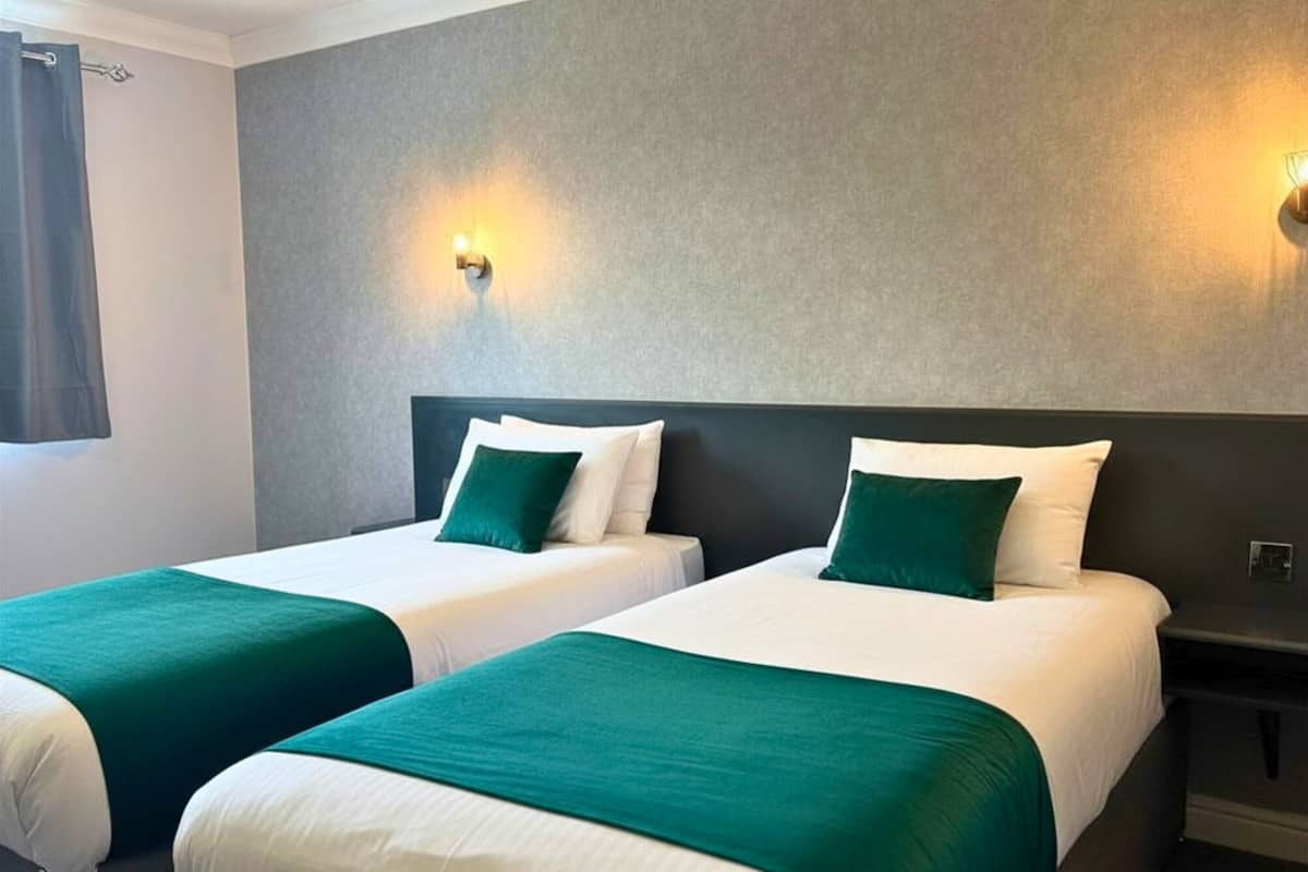 Quarto de hotel com duas camas, cada uma com colcha verde e roupa de cama branca, parede texturizada cinza e duas luminárias fixadas na parede.