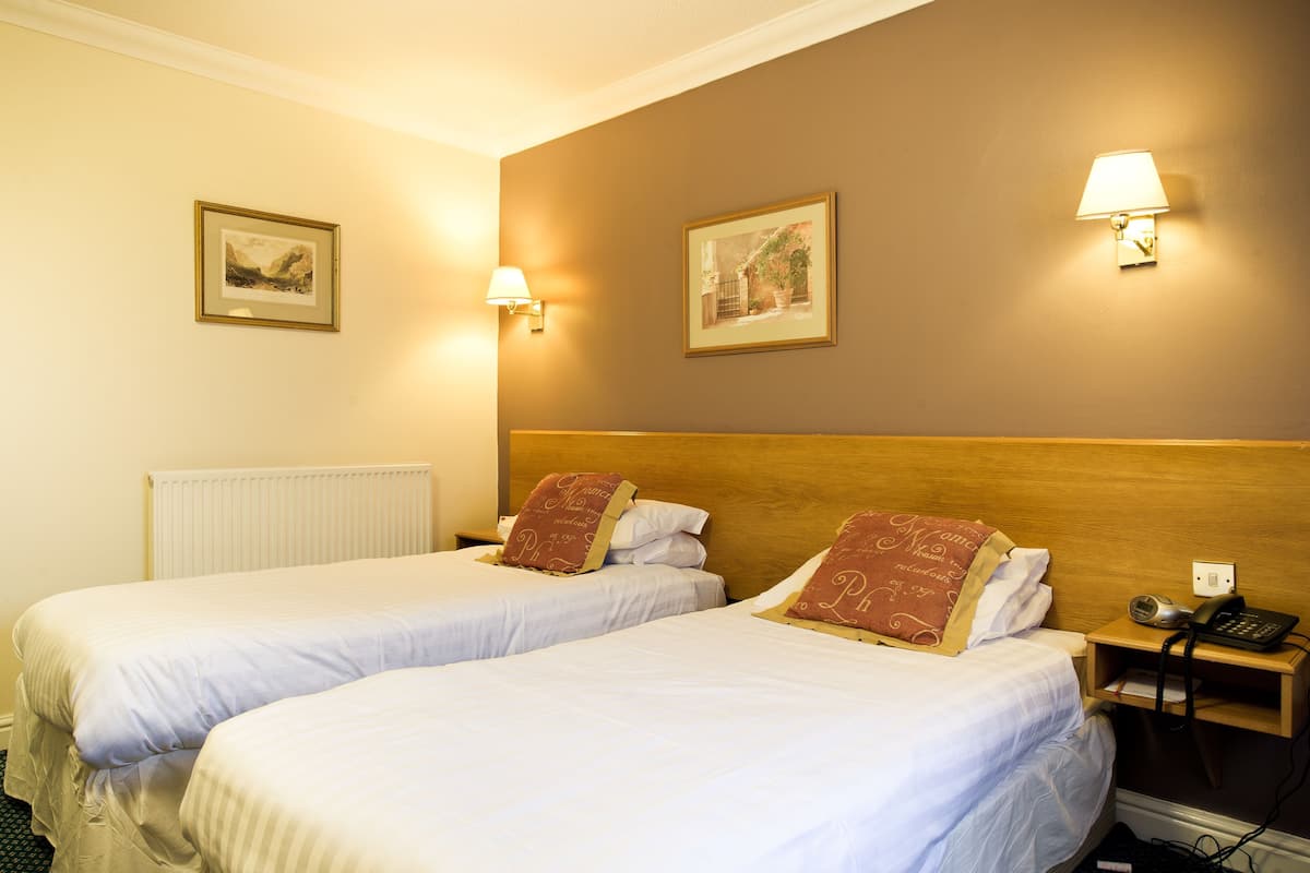 Quarto de hotel com duas camas, mesa de cabeceira com abajur, telefone e duas imagens emolduradas na parede.