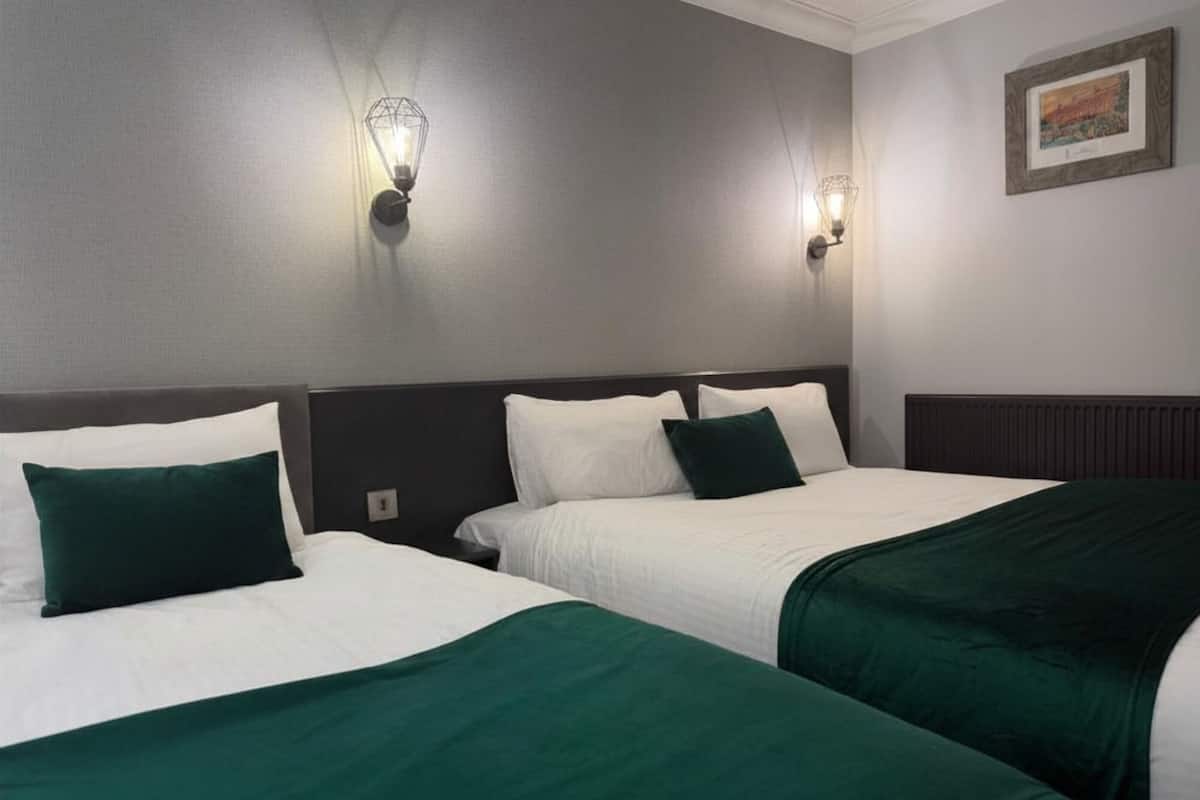 Quarto de hotel com duas camas, roupa de cama branca, almofadas decorativas verdes e luminárias fixadas na parede.