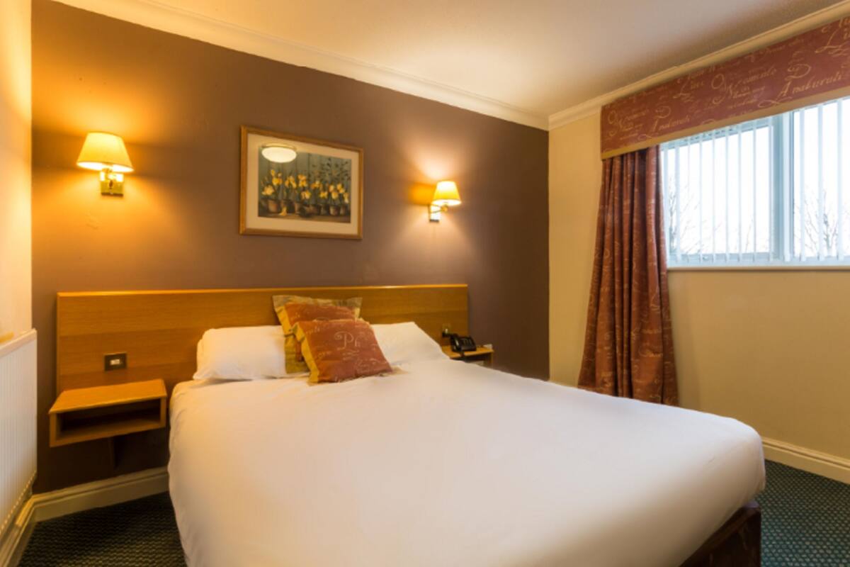 Quarto de hotel com uma cama grande, duas luminárias de cabeceira, uma mesa de apoio, um quadro na parede e uma janela com cortinas.