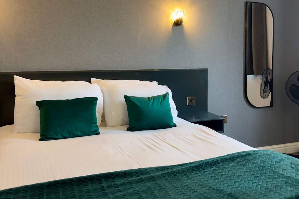 Cama bem arrumada com almofadas brancas e verdes, cabeceira escura, mesa de cabeceira e luminária fixada na parede.