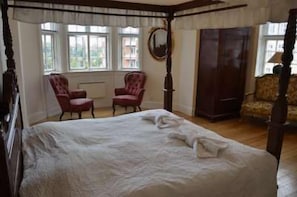 Uma cama com dossel, uma colcha branca, duas poltronas vermelhas, um guarda-roupa de madeira e um espelho na parede.