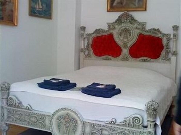 Uma cama com estrutura branca e detalhes entalhados, cabeceira estofada em vermelho e roupa de cama azul dobrada com cuidado.