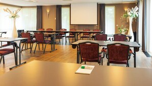 Sala de conferências com várias mesas e cadeiras, uma tela de projetor e plantas decorativas.