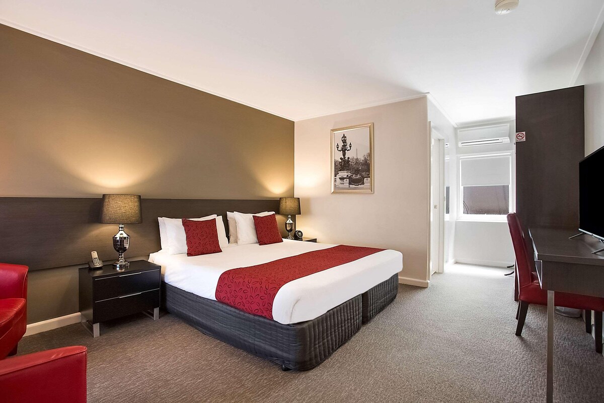 Quarto de hotel com cama grande, escrivaninha, cadeira, mesa de cabeceira e abajur.