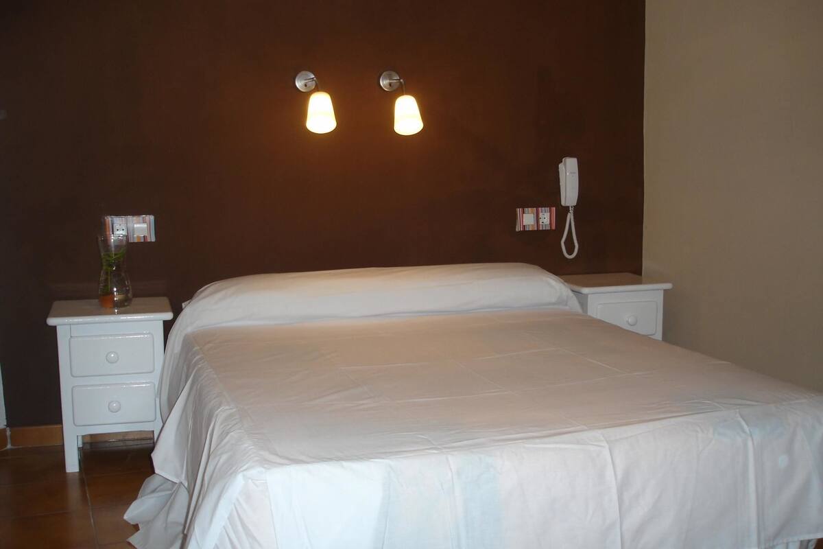Quarto de hotel com uma cama grande, mesa de cabeceira com telefone e luminárias fixadas na parede.
