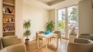 Uma sala de estar moderna com piso de madeira, uma mesa branca com um laptop, duas cadeiras, uma estante de livros e uma janela grande com vista para a cidade.