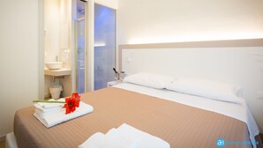 Quarto de hotel com cama, banheiro com pia e espelho, e mesa de cabeceira com abajur.