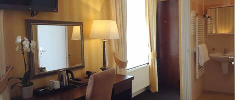 Quarto de hotel com escrivaninha, cadeira, luminária, espelho e vista para o banheiro.