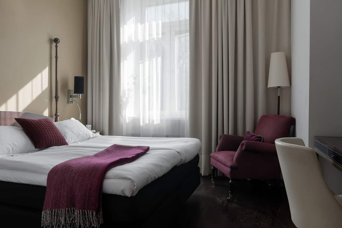 Quarto de hotel com cama, cadeira, luminária e janela com cortinas.