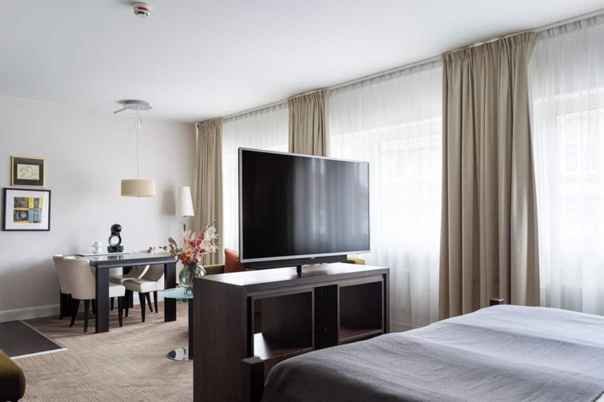 Quarto de hotel com cama, televisão de tela plana em um suporte, escrivaninha com cadeiras e área de jantar.
