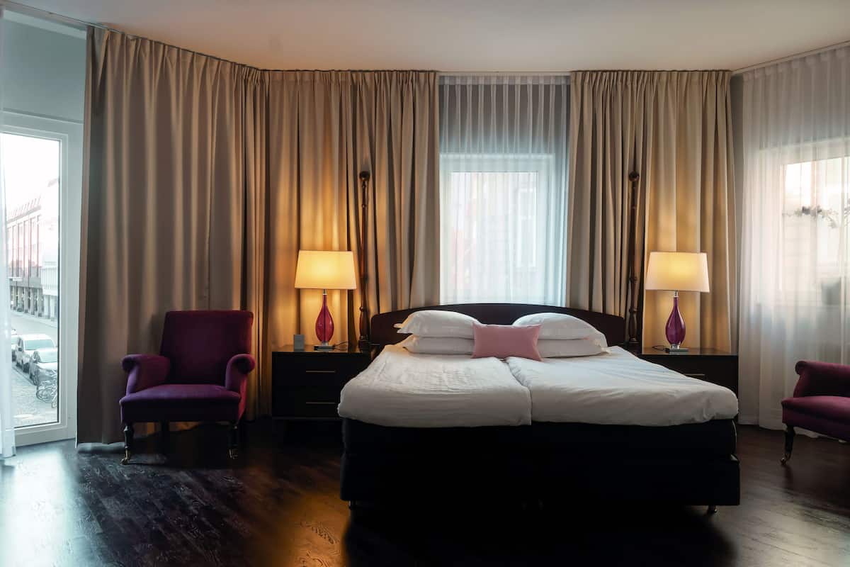 Quarto de hotel com uma cama grande, duas mesas de cabeceira com abajures, uma poltrona roxa e vista da rua por uma janela ampla.
