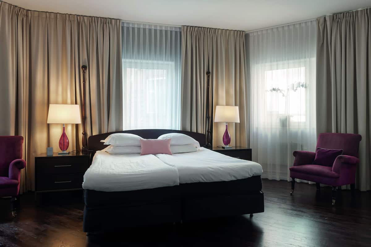 Quarto de hotel com uma cama grande, duas mesas de cabeceira com abajures, uma cadeira e uma janela com cortinas.