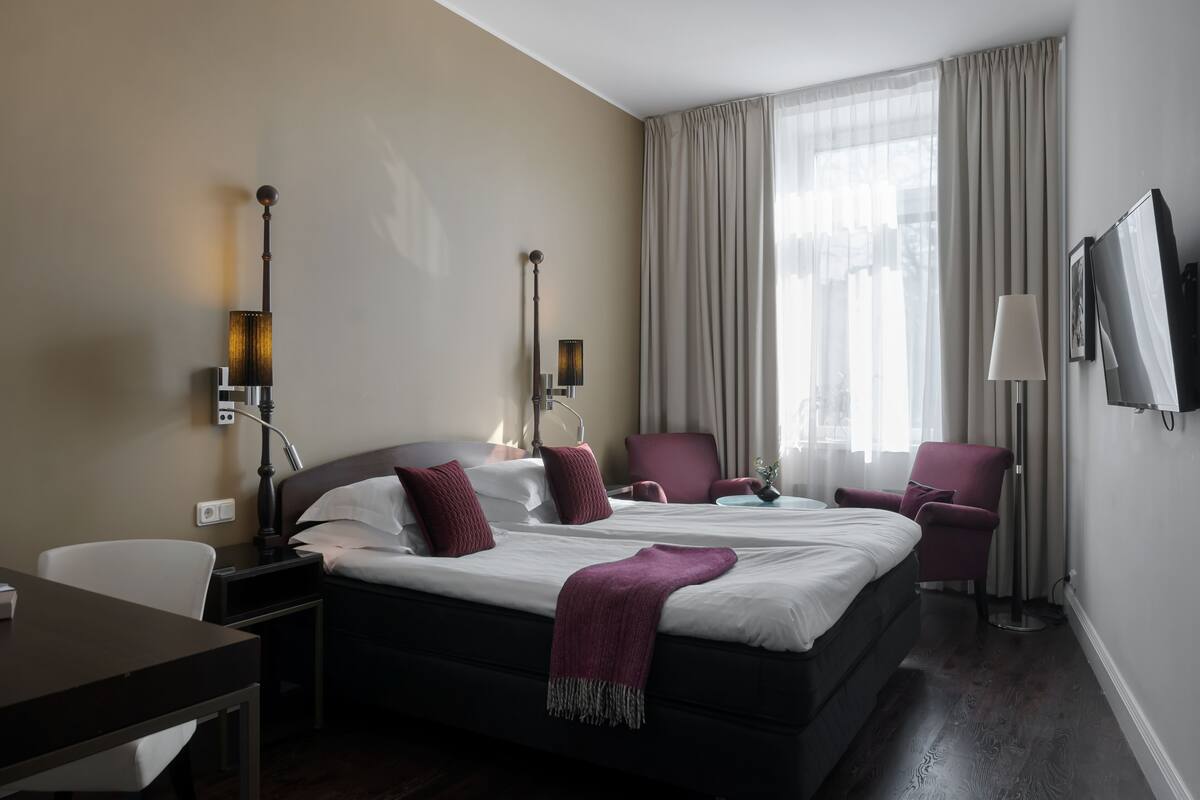 Quarto de hotel com uma cama grande, duas mesas de cabeceira, uma escrivaninha, uma cadeira, uma televisão e uma janela com cortinas.