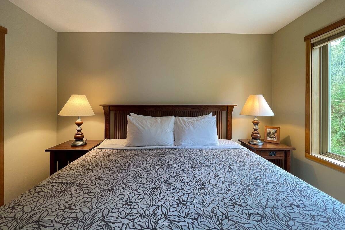 Cama bem arrumada com colcha estampada, acompanhada por duas mesas de cabeceira com abajures e quadros emoldurados.