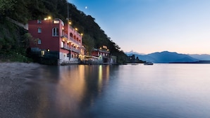 Um hotel à beira-mar com fachada vermelha, várias varandas e vista para o lago e montanhas ao fundo.