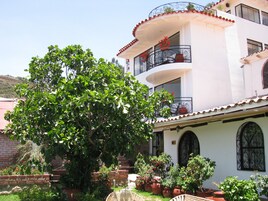 Um edifício de dois andares com varanda, plantas em vasos e uma grande árvore no pátio.
