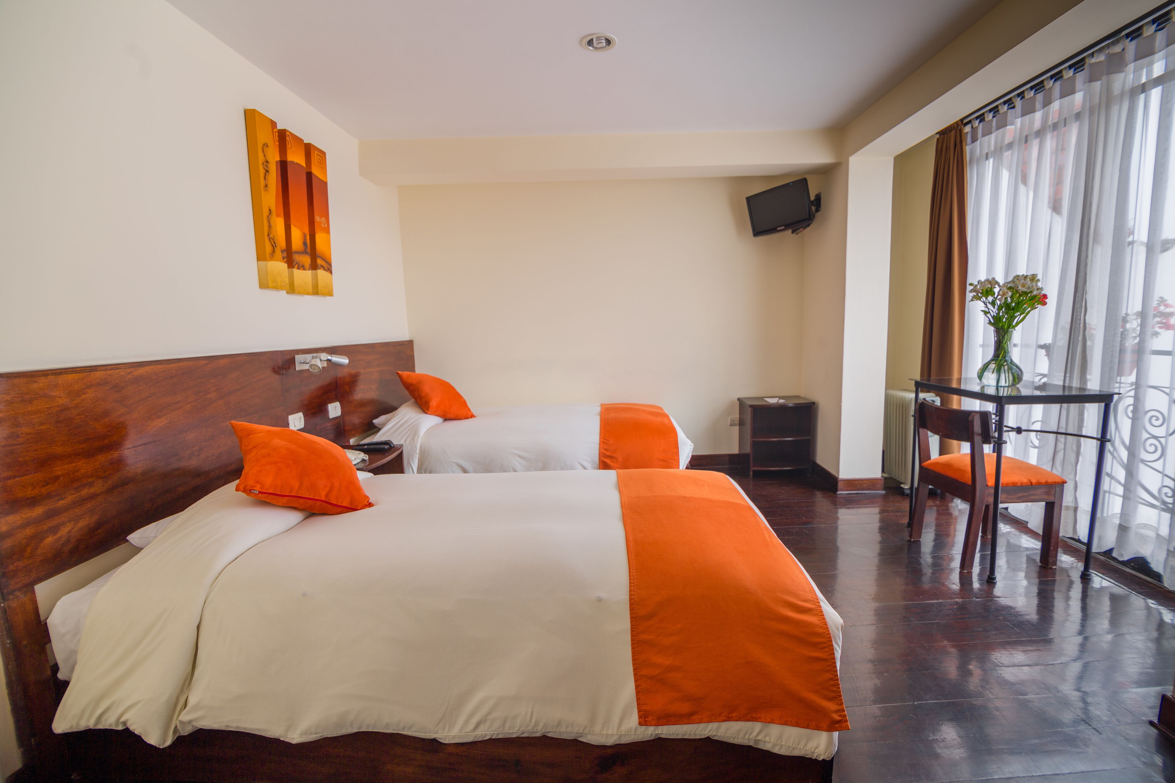 Quarto de hotel com duas camas, cabeceira de madeira, uma mesa pequena com uma cadeira, televisão fixada na parede e uma janela com cortinas.