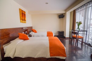 Quarto de hotel com duas camas, cabeceira de madeira, uma mesa pequena com uma cadeira, televisão fixada na parede e uma janela com cortinas.