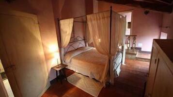 Uma cama de dossel com cortina, uma mesinha de cabeceira e um guarda-roupa em um quarto com piso de madeira e teto inclinado.