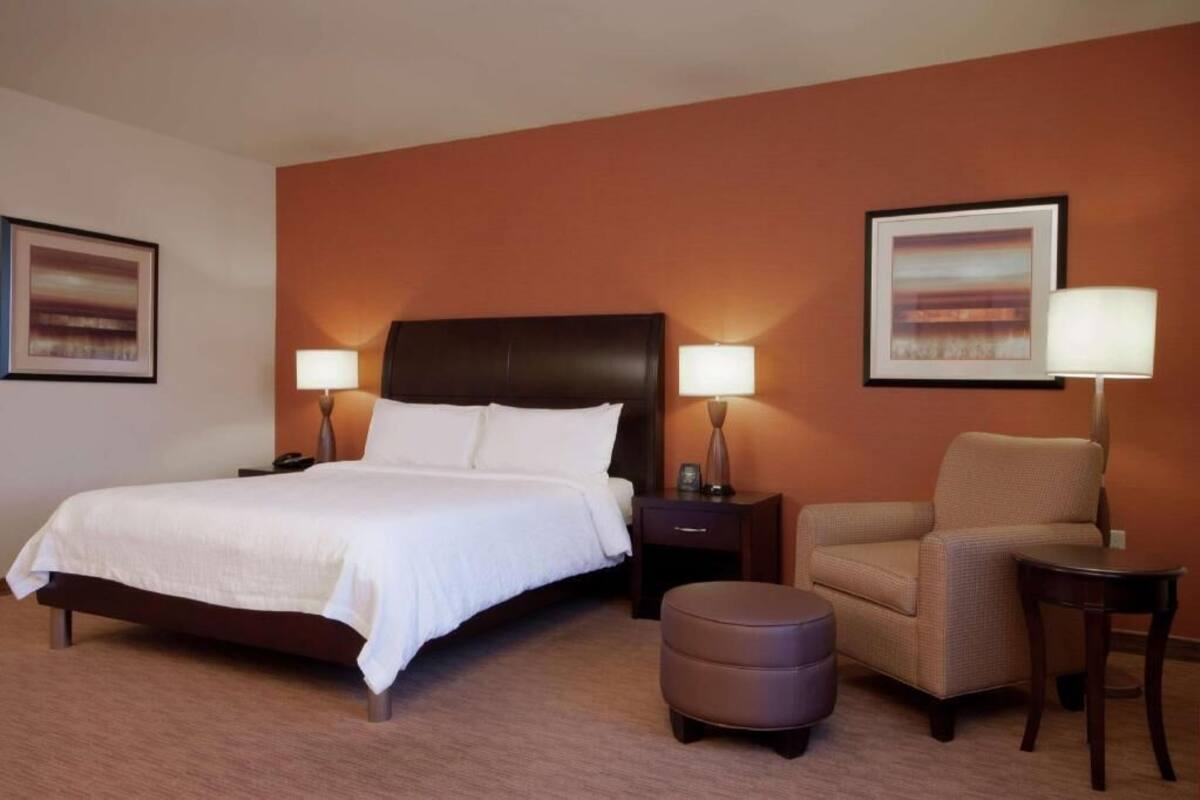 Quarto de hotel com uma cama, duas mesas de cabeceira, uma cadeira, um banquinho e duas luminárias fixadas na parede.