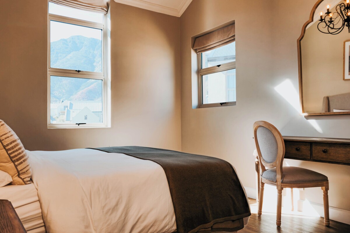 Quarto com cama, escrivaninha, cadeira e duas janelas com vista para montanhas.
