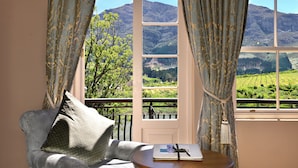 Quarto com vista para uma paisagem montanhosa através de uma grande janela, uma cadeira e uma pequena mesa com um livro.