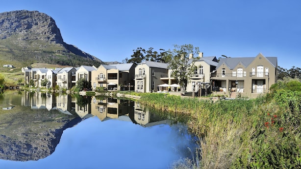 Um hotel na beira do lago, com vários edifícios refletidos na água, rodeado por vegetação e uma montanha ao fundo.