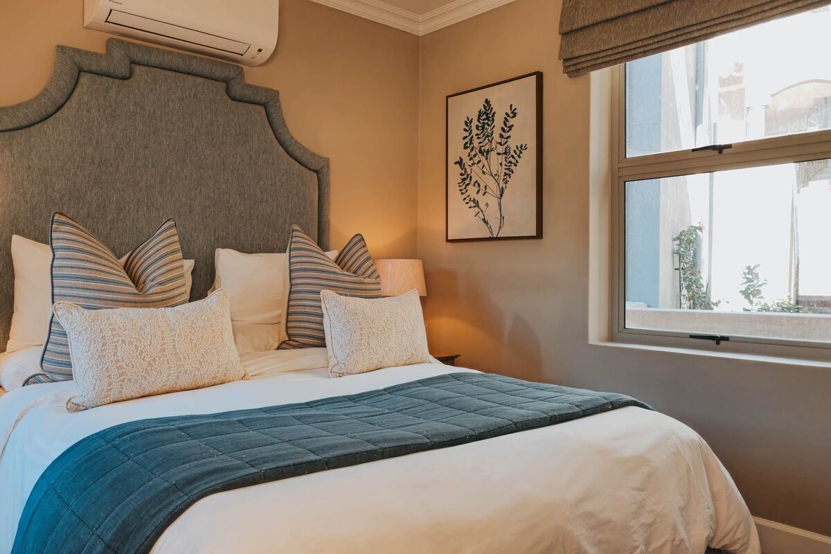 Cama bem arrumada com cabeceira, mesa de cabeceira com abajur e janela com vista para o exterior.