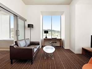 Quarto de hotel moderno com sofá de couro, uma pequena mesa redonda, uma escrivaninha com cadeira e grandes janelas com vista para a cidade.