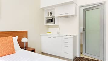 Quarto de hotel com cama, mesa de cabeceira, armário branco, micro-ondas, chaleira, pia e porta deslizante.