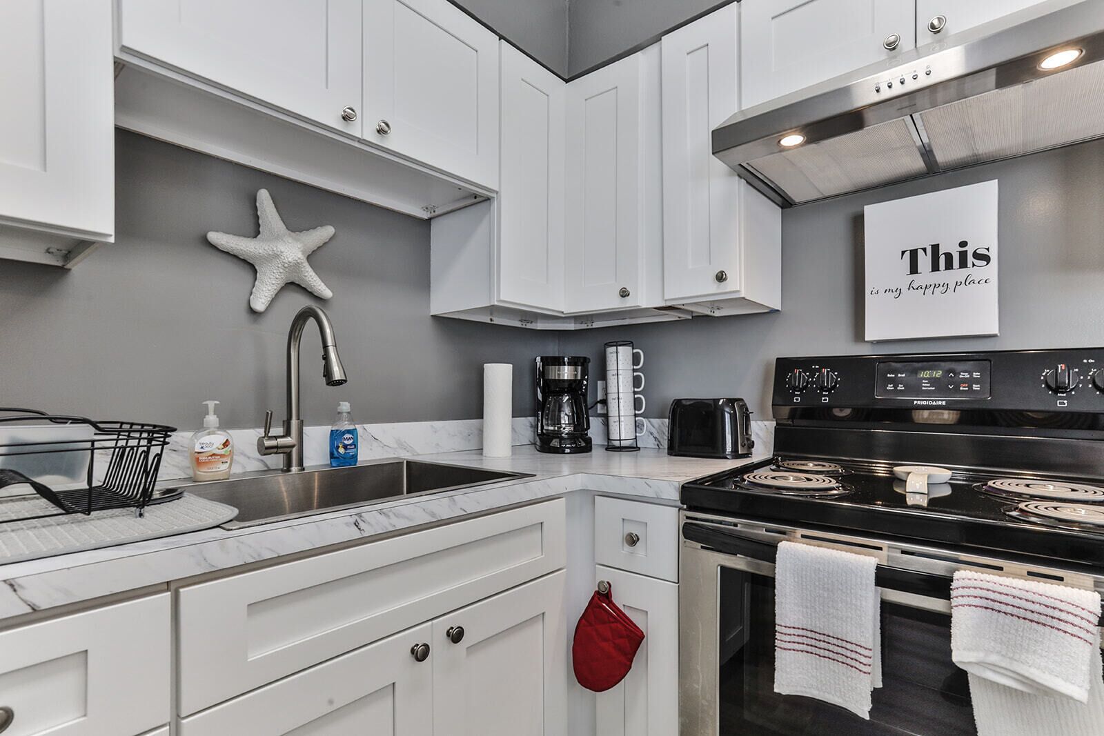 Cozinha moderna com armários brancos, fogão preto, decoração com estrela-do-mar e um letreiro que diz 'Este é o meu lugar feliz'.