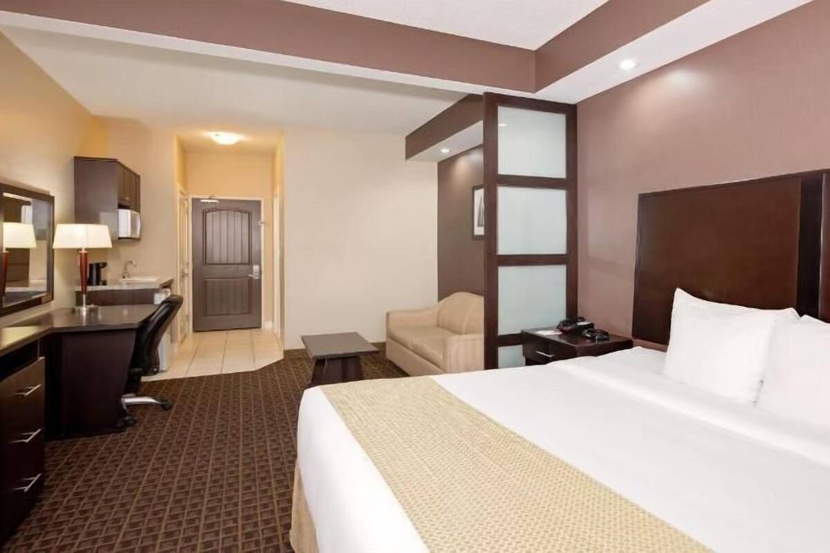 Quarto de hotel com uma cama grande, uma escrivaninha com cadeira, uma mesinha, um sofá e um banheiro visível através de uma porta de vidro.