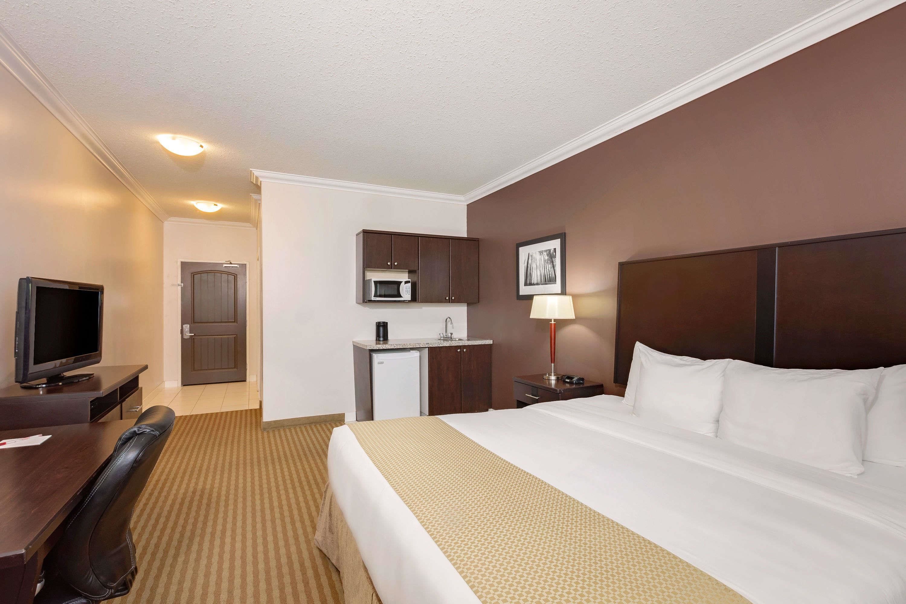 Quarto de hotel com cama, escrivaninha, cadeira, televisão, cozinha compacta e porta para o banheiro.